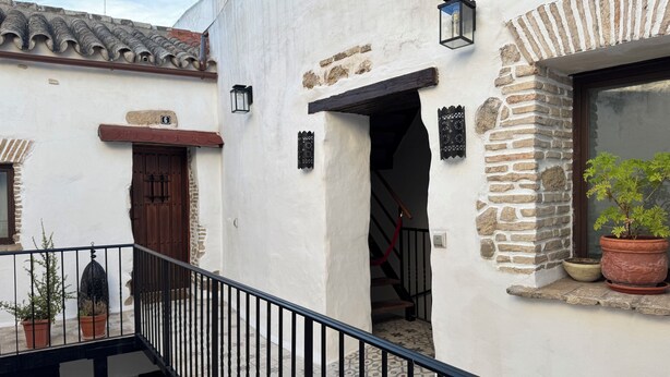 Um edifício tradicional com telhado de azulejos, porta de madeira e escadaria que leva a um andar superior. Há plantas em vasos na varanda e uma janela com vista para o céu.