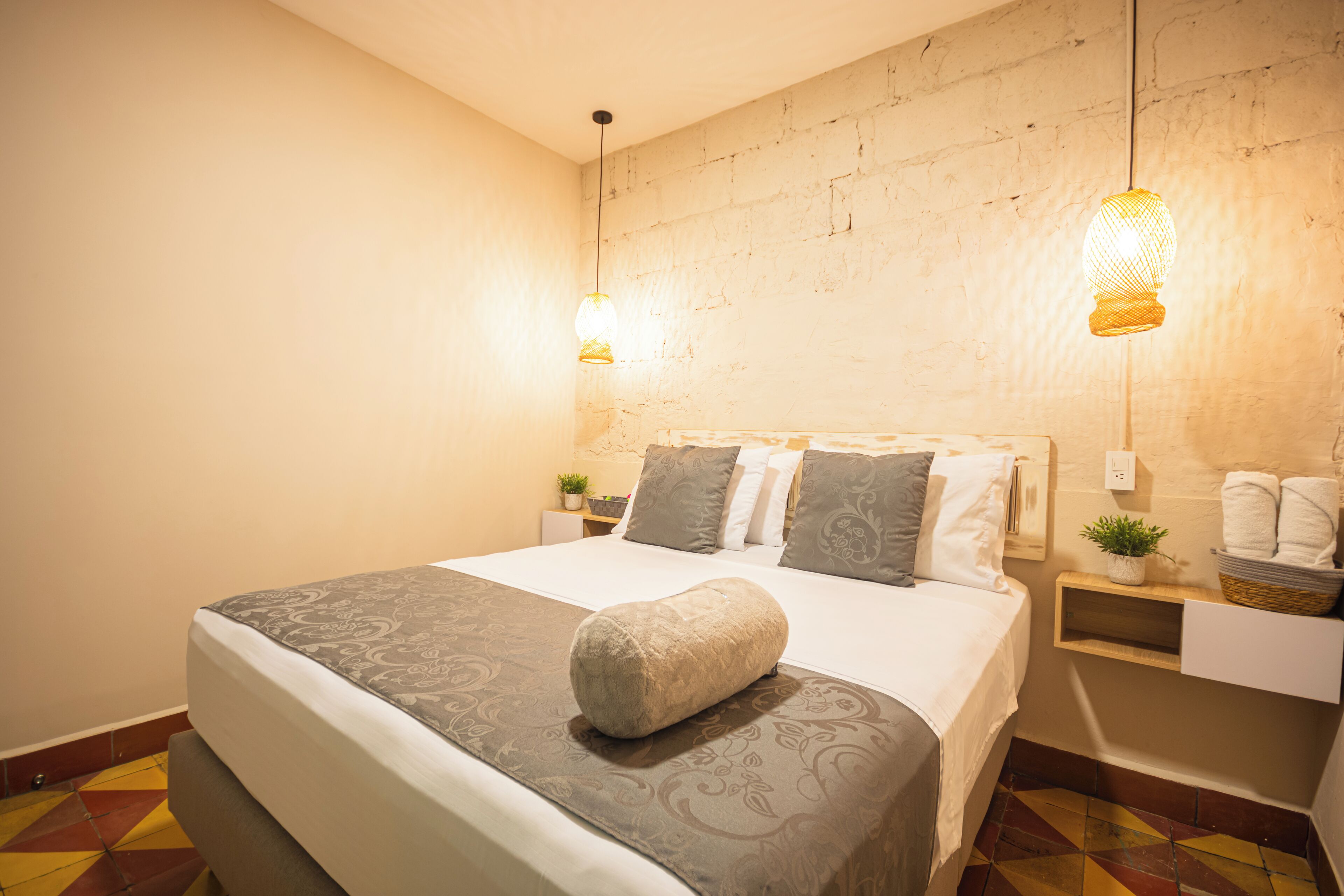 Cama bem arrumada com travesseiro decorativo, mesa de cabeceira com planta e prateleira na parede com toalhas.