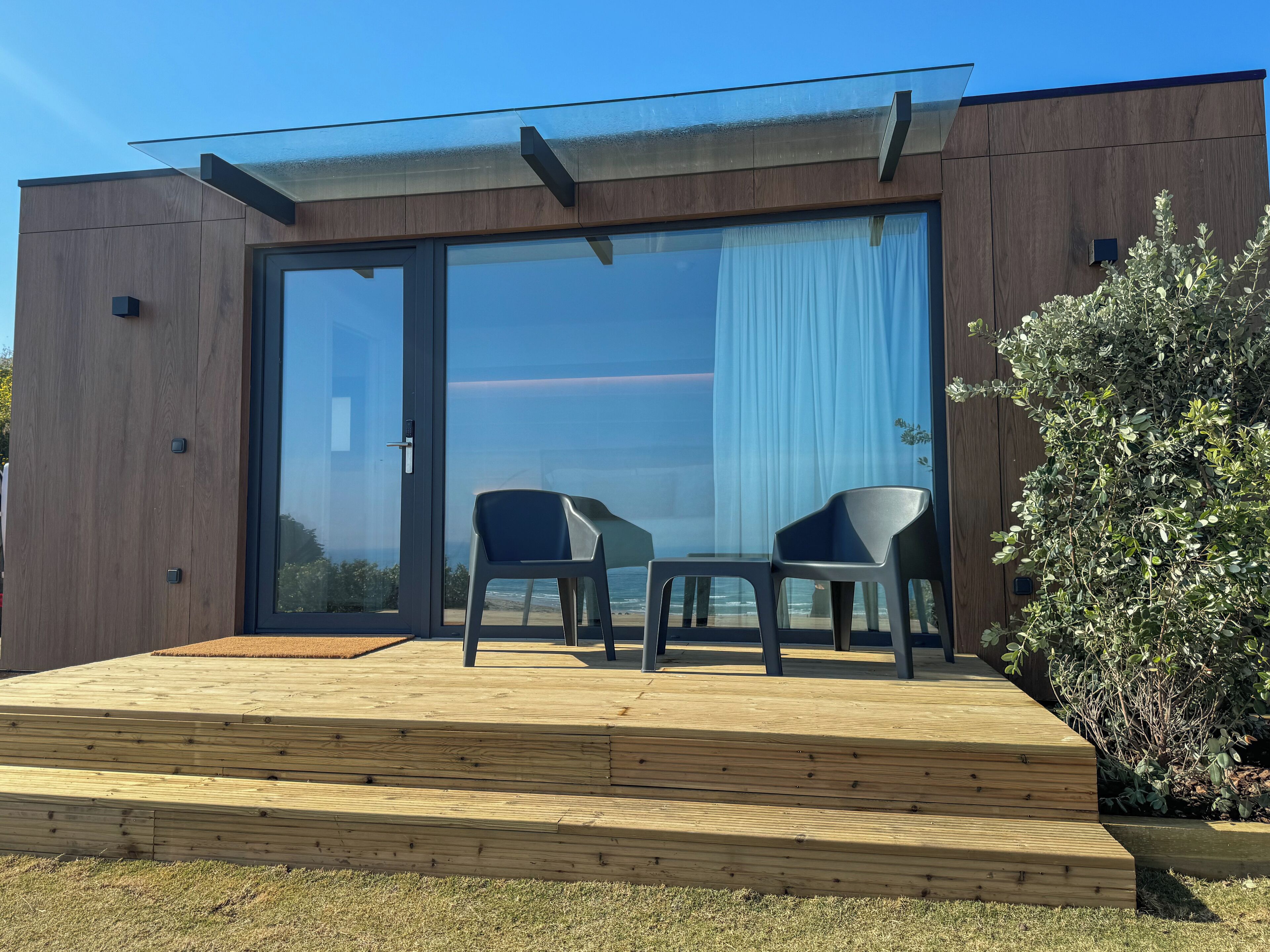 Um edifício moderno com porta de vidro que dá para uma área externa com assentos, incluindo duas cadeiras e uma mesa.