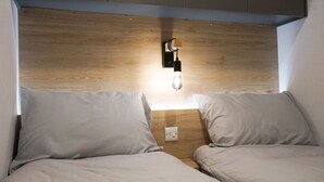 Quarto com cabeceira de madeira, duas camas com roupa de cama cinza e luminária fixada na parede.