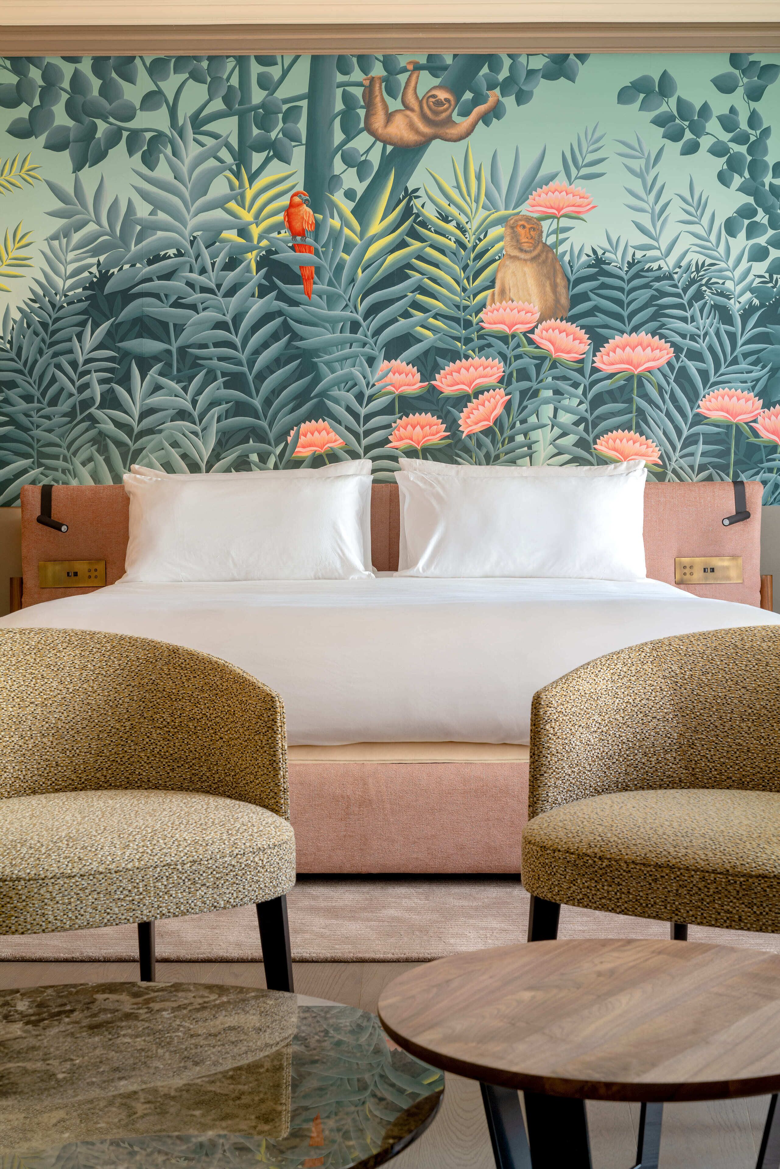 Quarto de hotel com cama, duas poltronas estofadas, uma mesinha redonda e um mural com um macaco e flores.