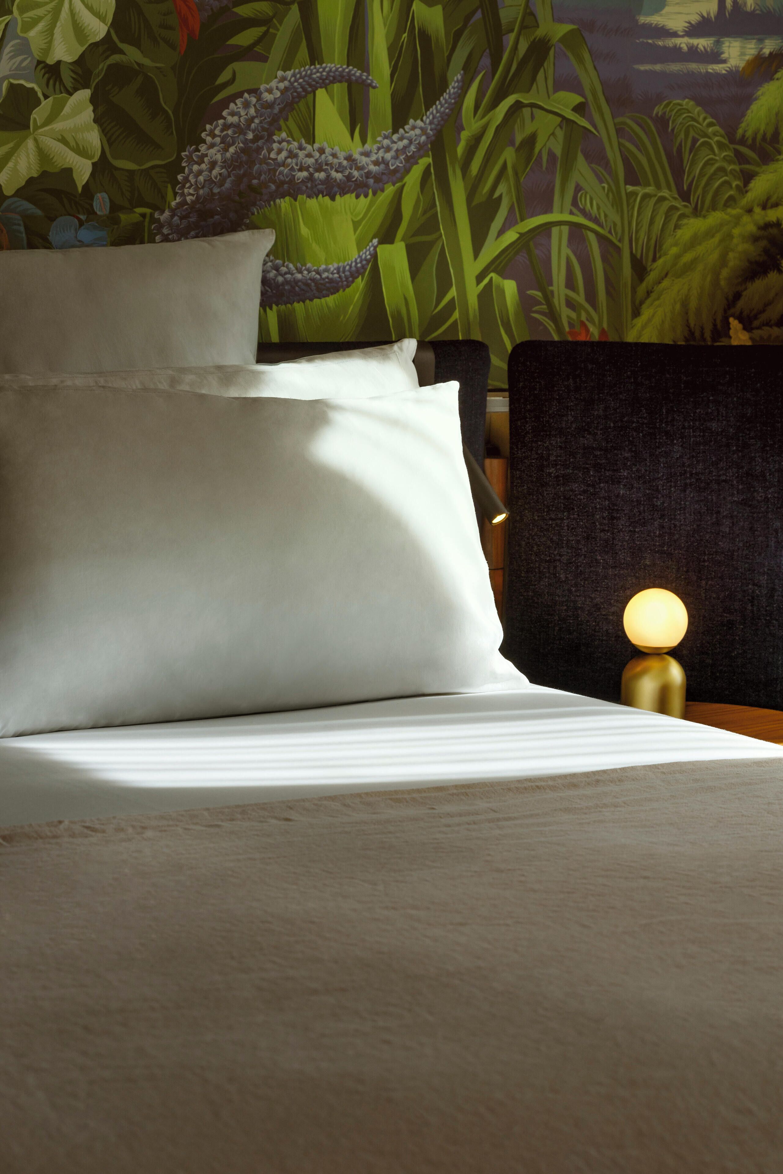 Uma cama com roupa de cama branca, um travesseiro branco e uma cadeira escura com uma luminária.