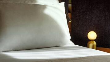 Uma cama com roupa de cama branca, um travesseiro branco e uma cadeira escura com uma luminária.