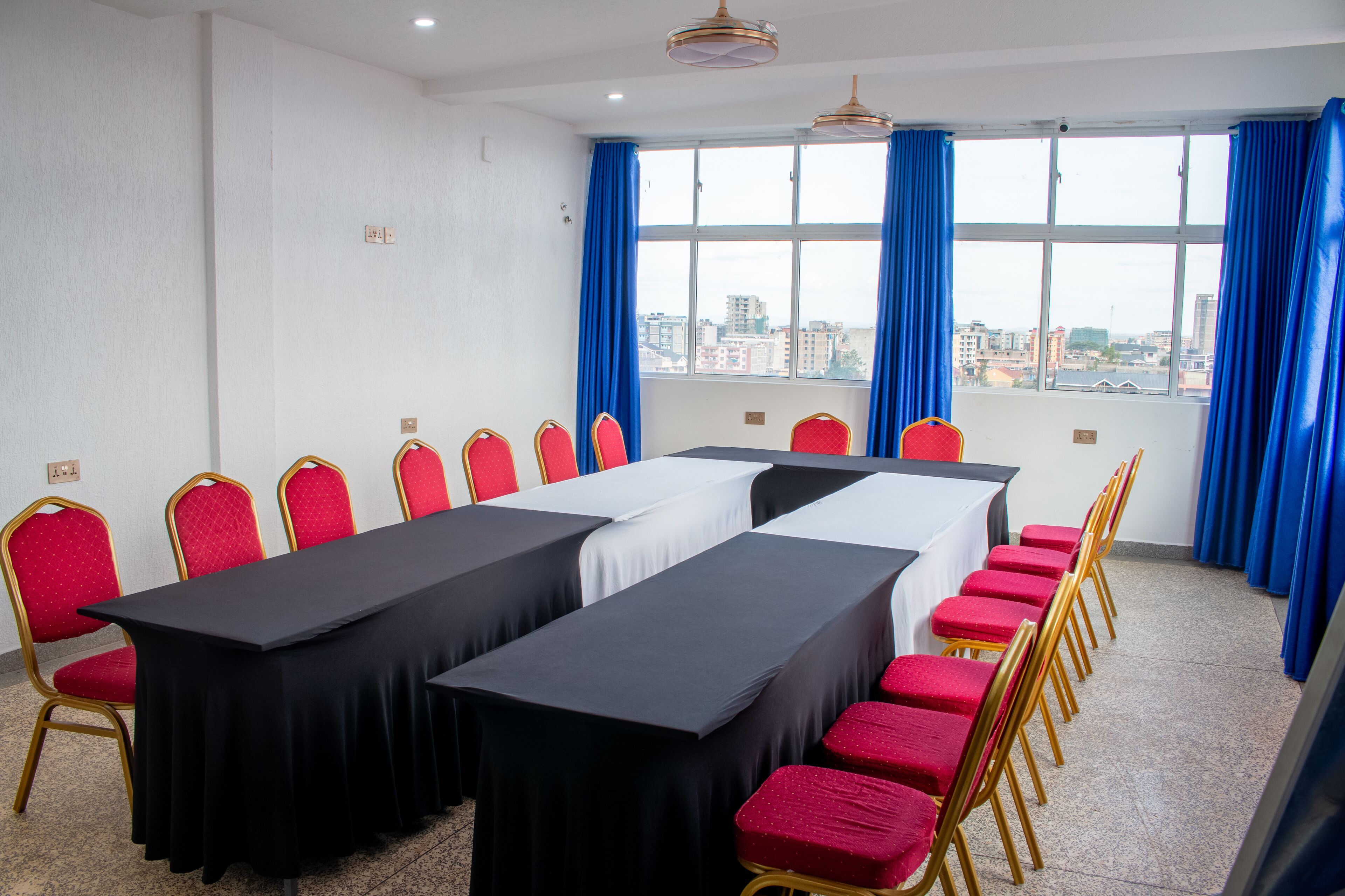 Uma sala de conferências com uma mesa comprida, cadeiras vermelhas e douradas, e grandes janelas com cortinas azuis.