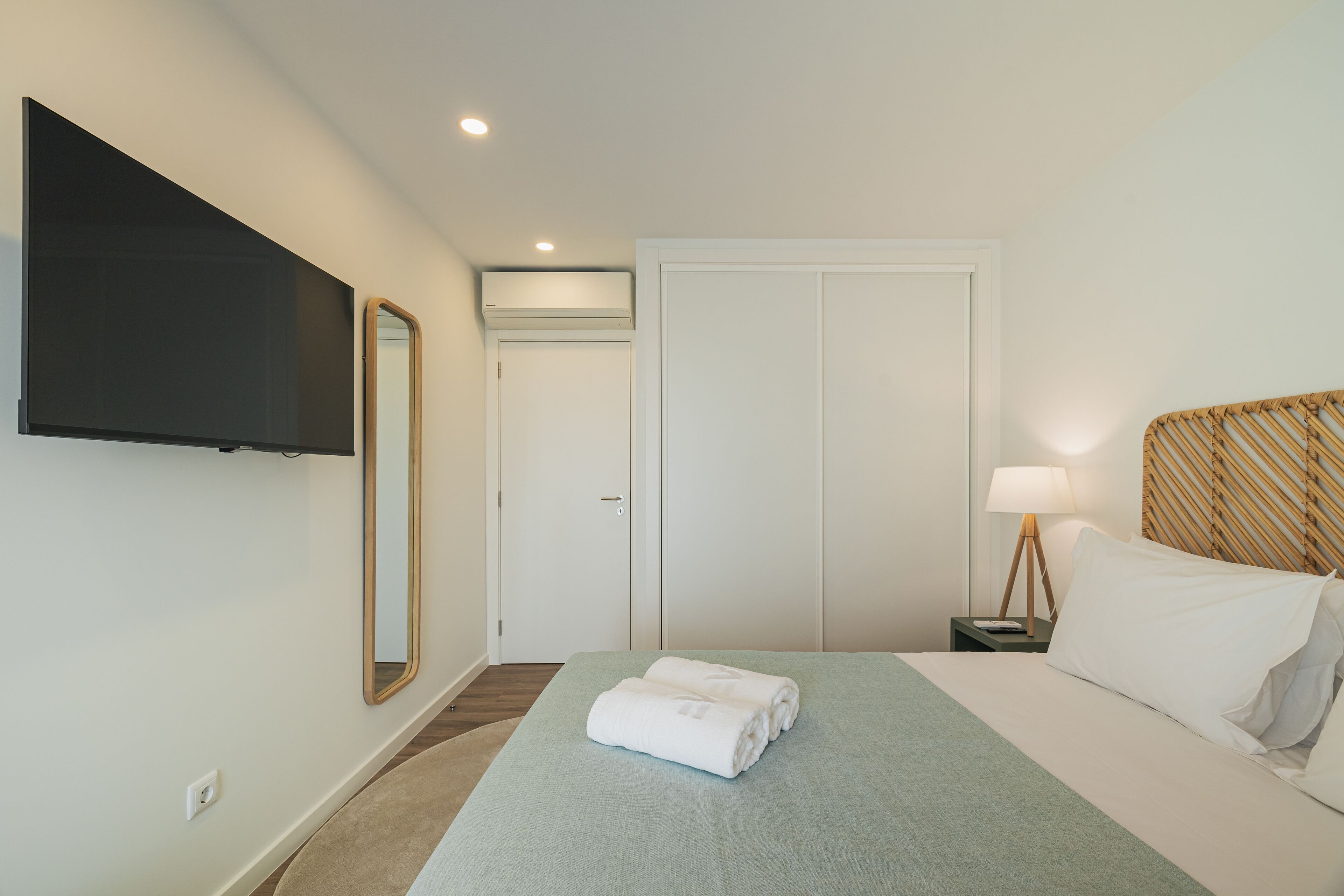 Quarto de hotel com cama, televisão fixada na parede, mesa de cabeceira com abajur e guarda-roupa.