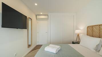 Quarto de hotel com cama, televisão fixada na parede, mesa de cabeceira com abajur e guarda-roupa.