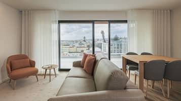 Uma sala de estar moderna com sofá, poltrona e mesa de jantar, janelas amplas com cortinas e vista para a cidade.