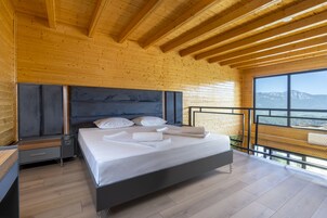 Quarto moderno com uma cama grande, vigas de madeira no teto, vista para montanhas através de uma varanda e uma mesa de cabeceira com abajur.