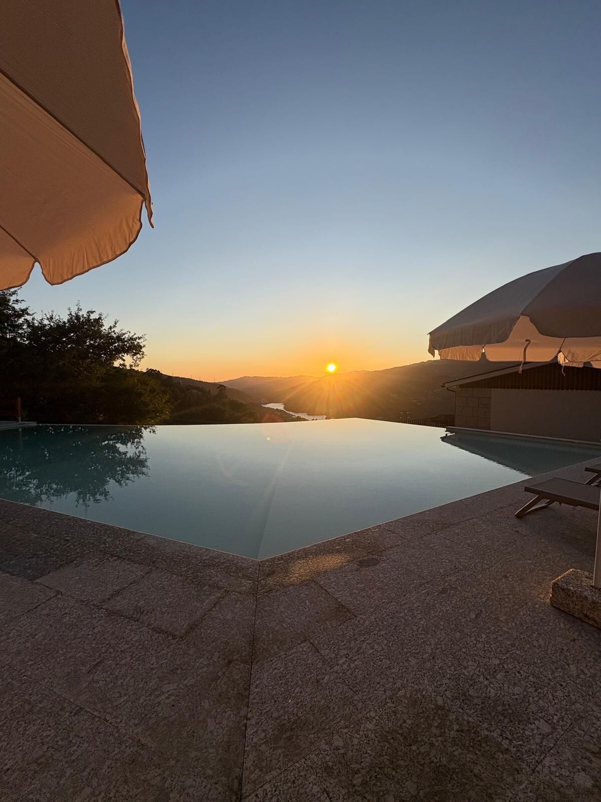 Piscina com vista para o pôr do sol, cadeiras de sol e guarda-sóis.