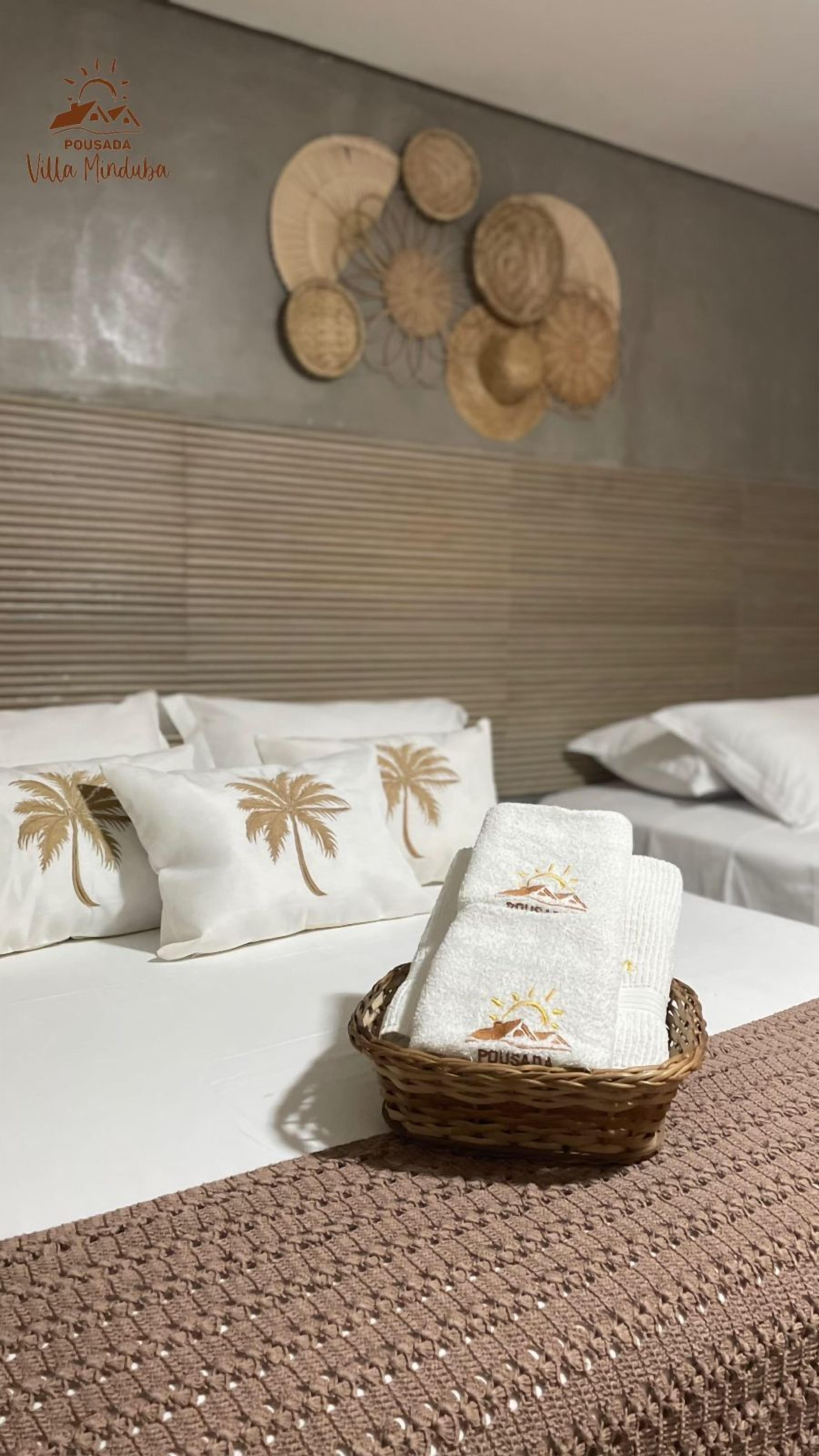 Cama bem arrumada com roupa de cama branca e almofadas de palmeira, um cesto de palha com toalhas e uma parede com um desenho circular.