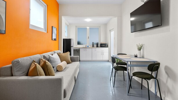 Uma sala de estar moderna com sofá cinzento, paredes laranja, uma televisão de tela plana e uma cozinha ao fundo.