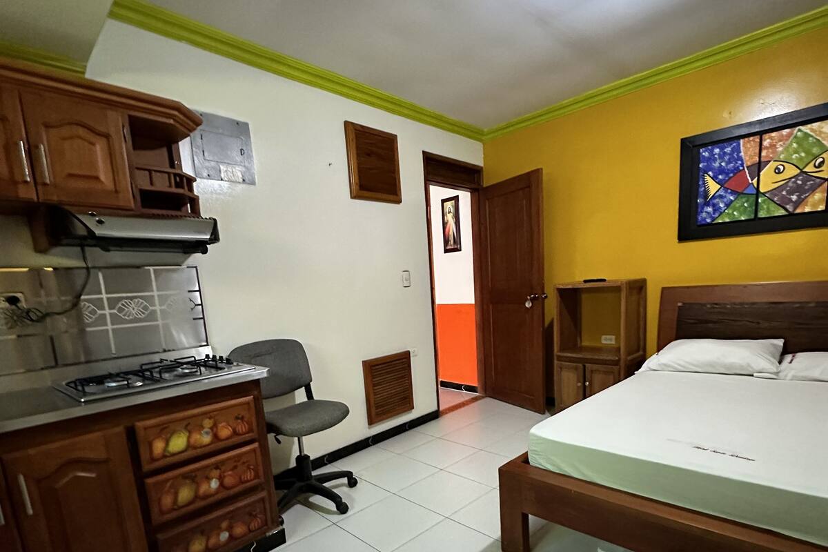 Um quarto com uma cama, uma escrivaninha com cadeira, um fogão e uma porta que dá para outro quarto.