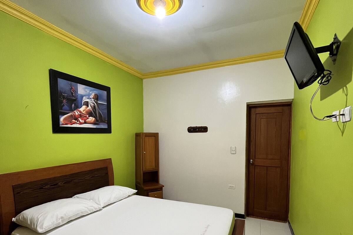 Quarto com cama, mesa de cabeceira, TV fixada na parede, porta e um quadro na parede.