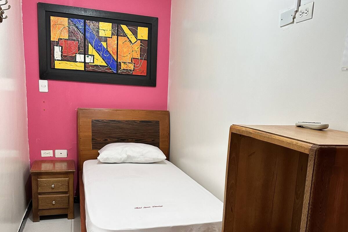 Quarto com cama, mesa de cabeceira, guarda-roupa de madeira e um quadro abstrato colorido na parede.