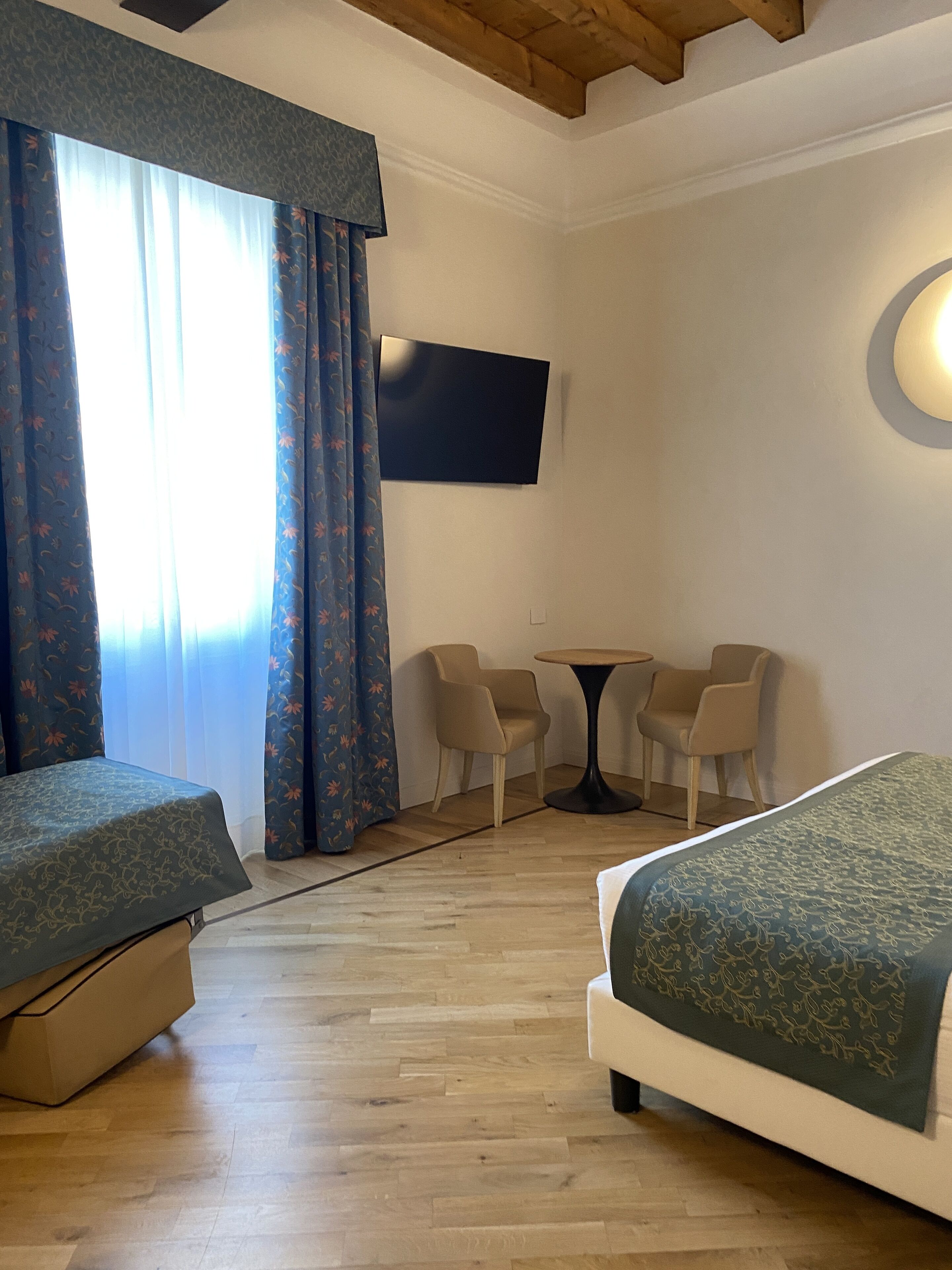 Quarto de hotel com cama, sofá, mesa pequena, duas cadeiras, TV na parede e janela com cortinas.
