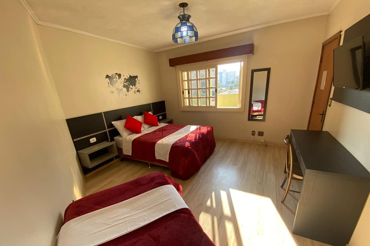 Quarto de hotel com duas camas, uma escrivaninha, cômoda, televisão e janela com vista.