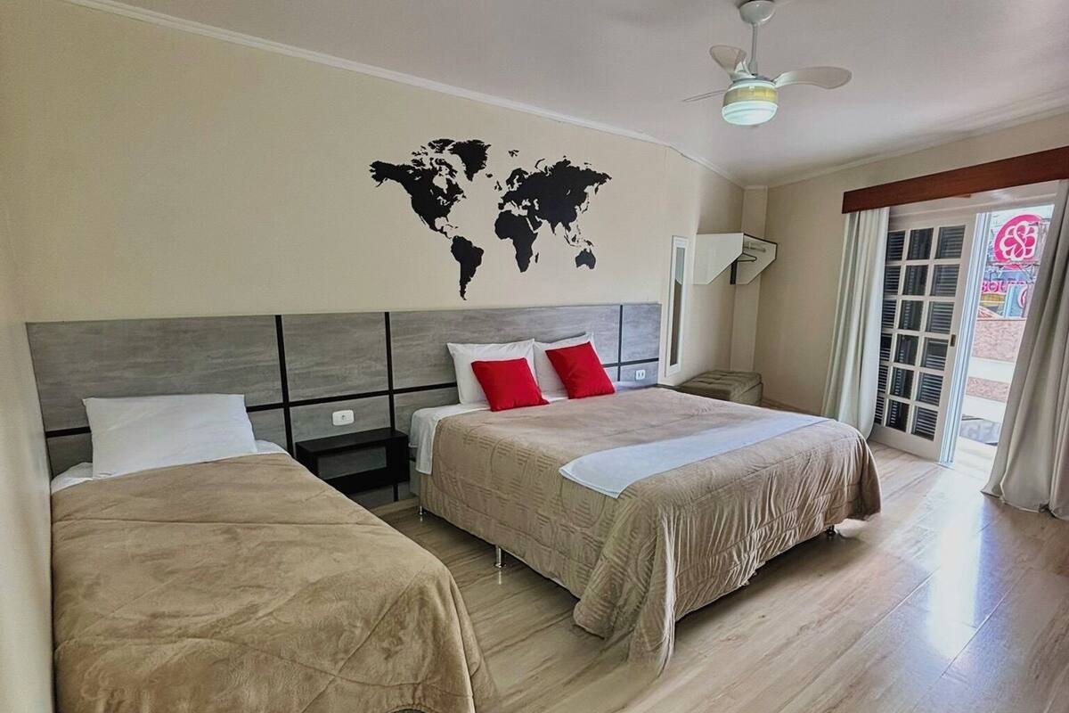 Quarto de hotel com duas camas, um mapa-múndi na parede, um ventilador de teto e vista para o exterior através de uma janela.