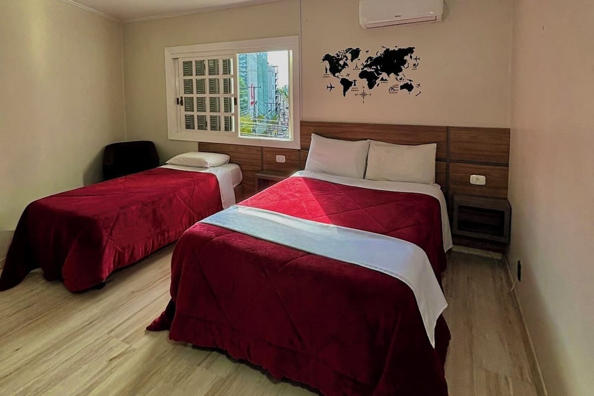 Quarto de hotel com duas camas, um ventilador de teto, um aparelho de ar condicionado, uma janela com vista para edifícios e um mapa-múndi na parede.