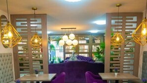 Restaurante com cadeiras roxas e mesas de madeira, uma área de estar central com sofá roxo e luminárias pendentes.