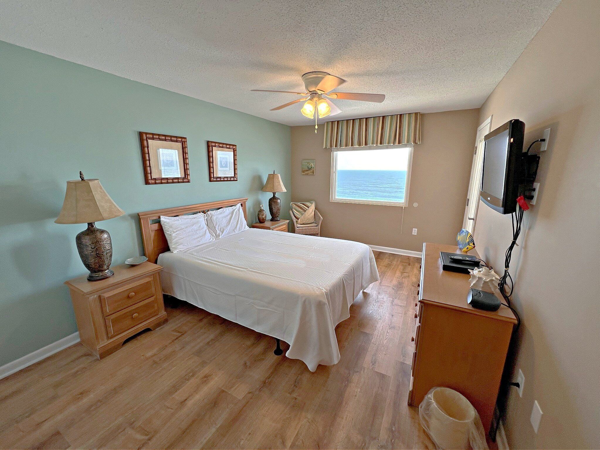 Quarto com cama, mesas de cabeceira, ventoinha no teto, janela com vista e televisão fixada na parede.