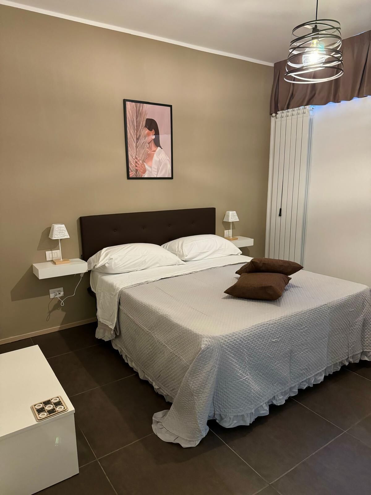 Quarto com cama, duas mesas de cabeceira, um criado-mudo, um quadro na parede e uma janela com cortinas.