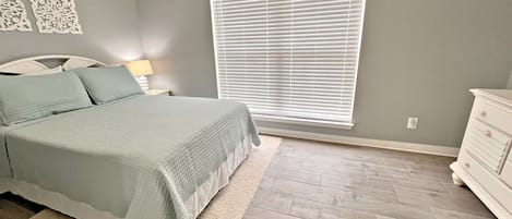 Um quarto com cama, mesa de cabeceira, cômoda, janela com persianas e painéis decorativos na parede.