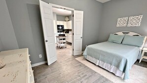 Um quarto com cama, mesa de cabeceira, cômoda e porta que dá para uma cozinha.
