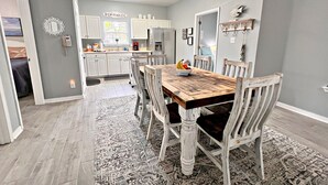Uma área de refeições com mesa de madeira e cadeiras brancas, uma cozinha ao fundo e um tapete estampado no chão.