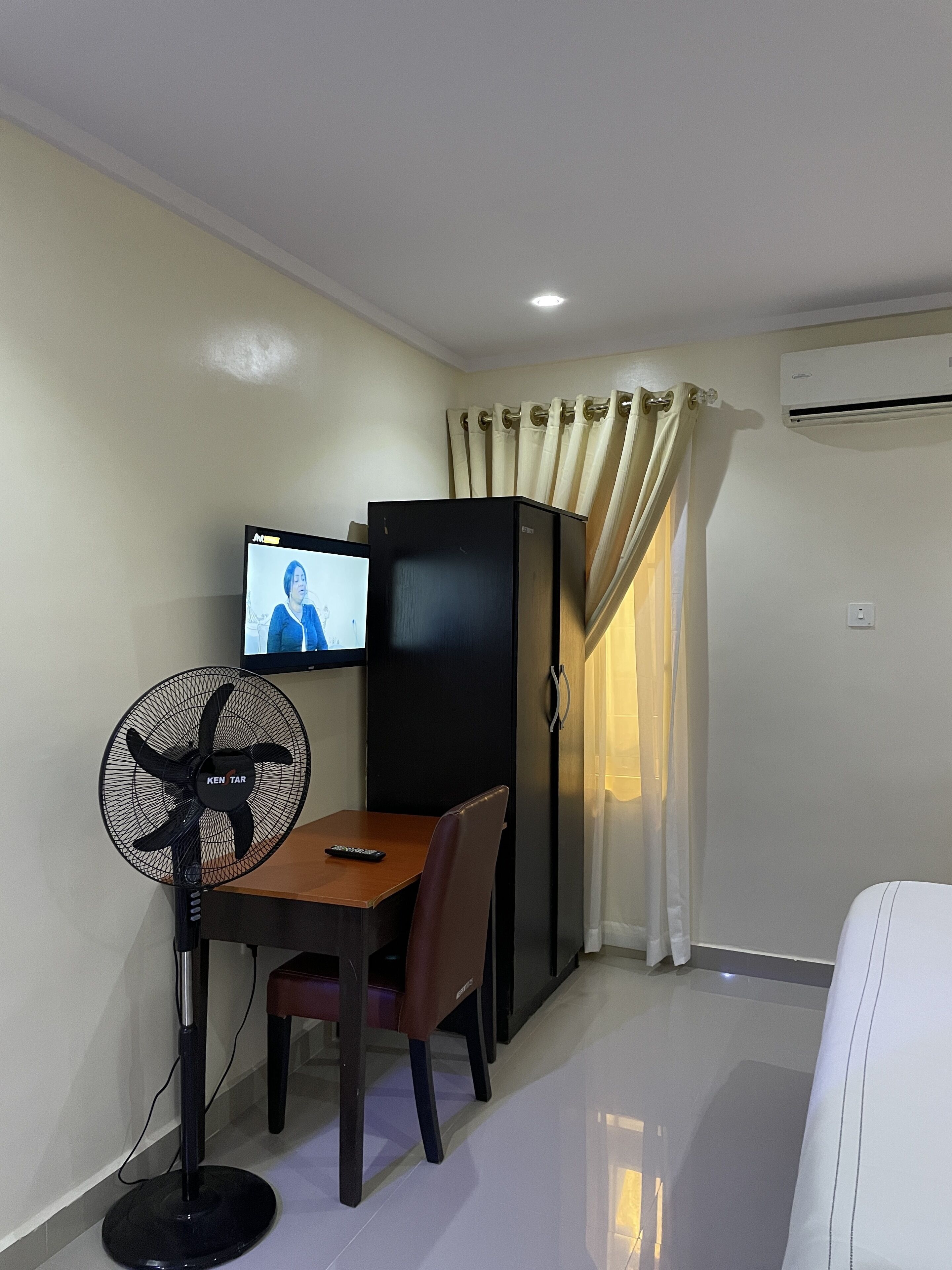 Quarto compacto com televisão, geladeira preta, mesa de jantar com cadeiras, ventilador e sofá branco.