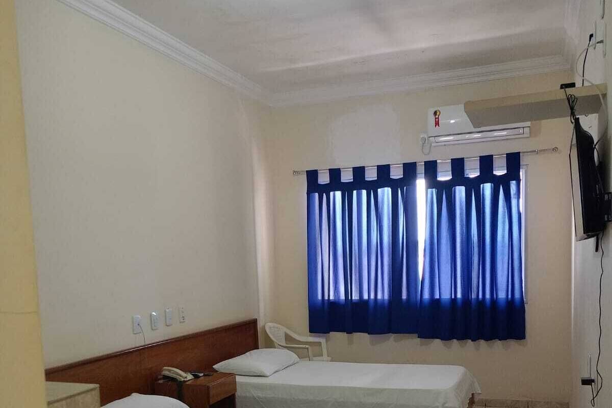 Quarto de hotel com duas camas, uma secretária pequena, uma cadeira, televisão fixada na parede e uma janela com cortinas azuis.