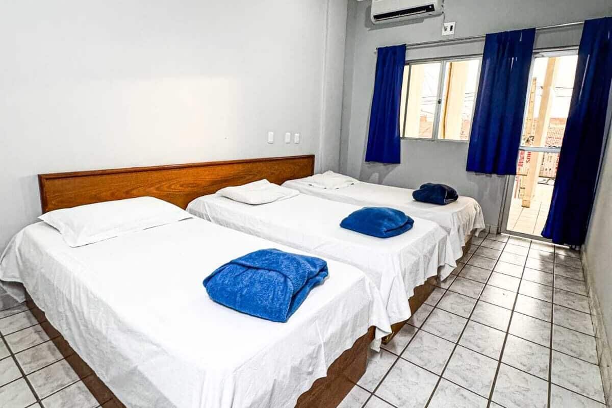 Quarto de hotel com três camas, roupa de cama branca, almofadas azuis e chão de azulejos.