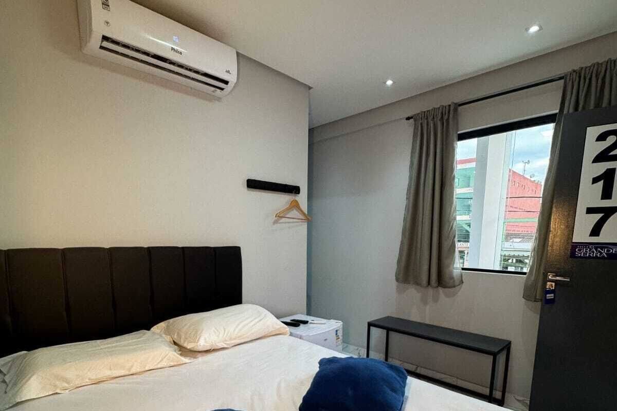 Quarto de hotel com duas camas, ar condicionado, janela com cortinas e vista para um edifício.