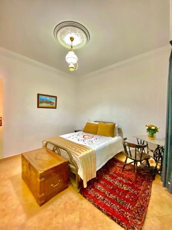 Quarto com cama, escrivaninha, cadeira, uma pequena mesa com um vaso e um tapete estampado vermelho.