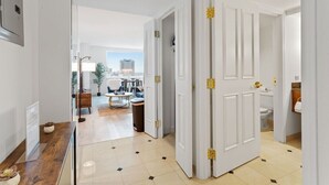 Corredor com balcão de madeira, porta branca com dobradiças douradas e vista para uma sala de estar com paisagem urbana.