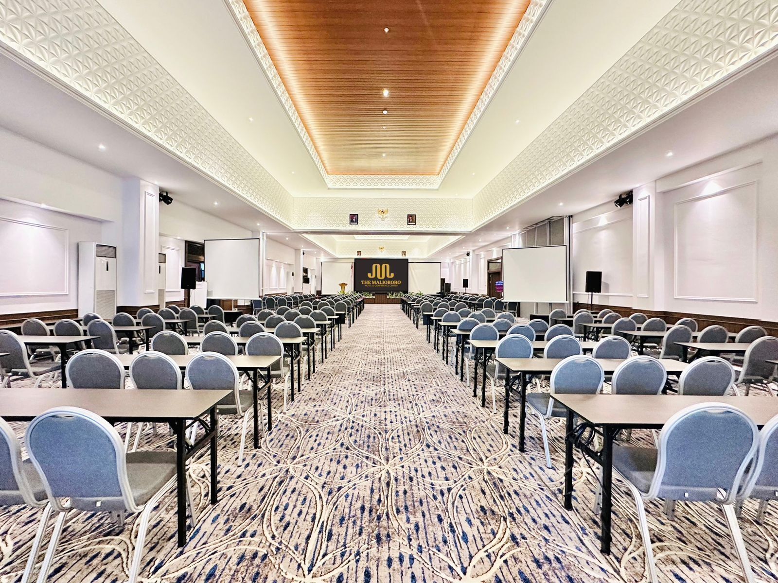 Uma sala de conferências com mesas e cadeiras dispostas em fileiras, um tapete estampado e teto de madeira.