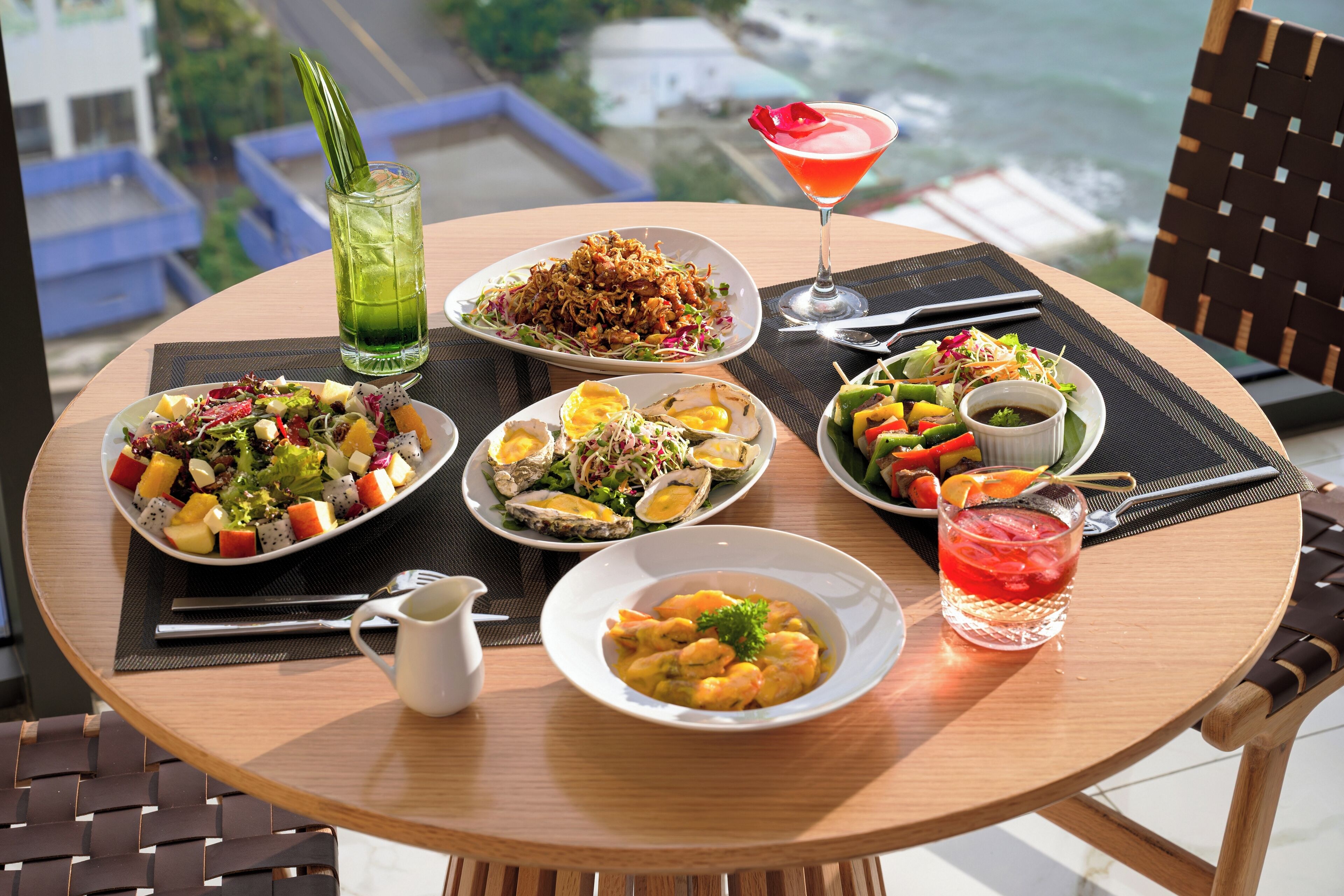 Uma mesa com vários pratos e bebidas, vista para o oceano e paisagem urbana ao fundo.