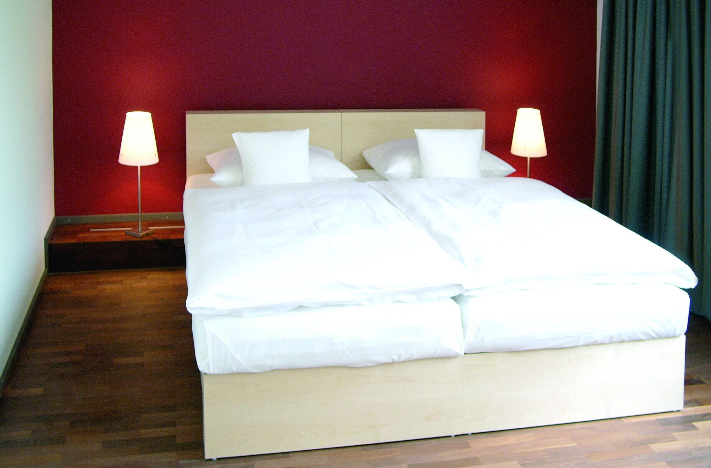 Uma cama bem arrumada com lençóis e almofadas brancas, duas luminárias de cabeceira e uma cabeceira de madeira.