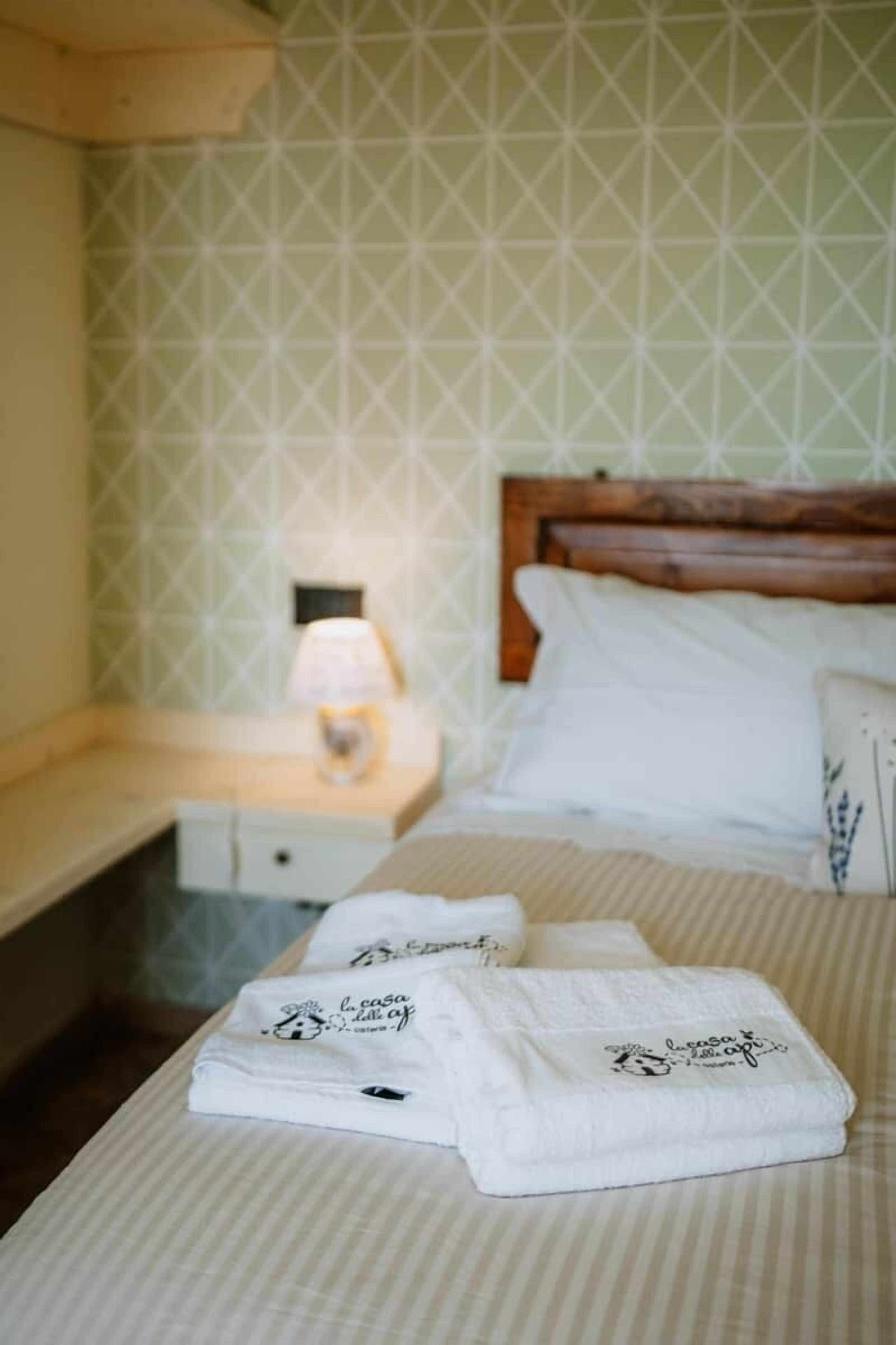 Uma cama com lençóis brancos e cabeceira, uma mesinha de cabeceira com abajur, e uma parede com padrão.