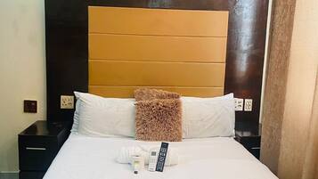 Quarto de hotel com uma cama grande, cabeceira de madeira, roupa de cama branca, almofada marrom e um controle remoto, além de uma garrafa de loção.