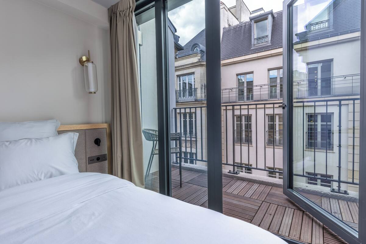 Quarto de hotel com uma janela grande, uma cama e vista para edifícios vizinhos.