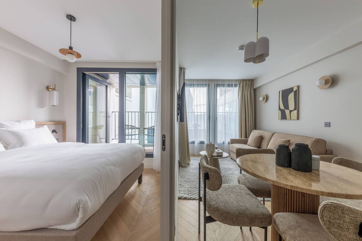 Quarto de hotel moderno com uma cama grande, área de jantar com mesa redonda e cadeiras, e um sofá.