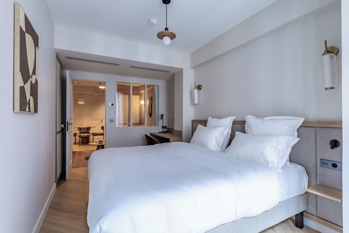 Quarto de hotel moderno com uma cama grande, mesas de cabeceira, uma escrivaninha e uma zona de refeições visível através de uma porta aberta.