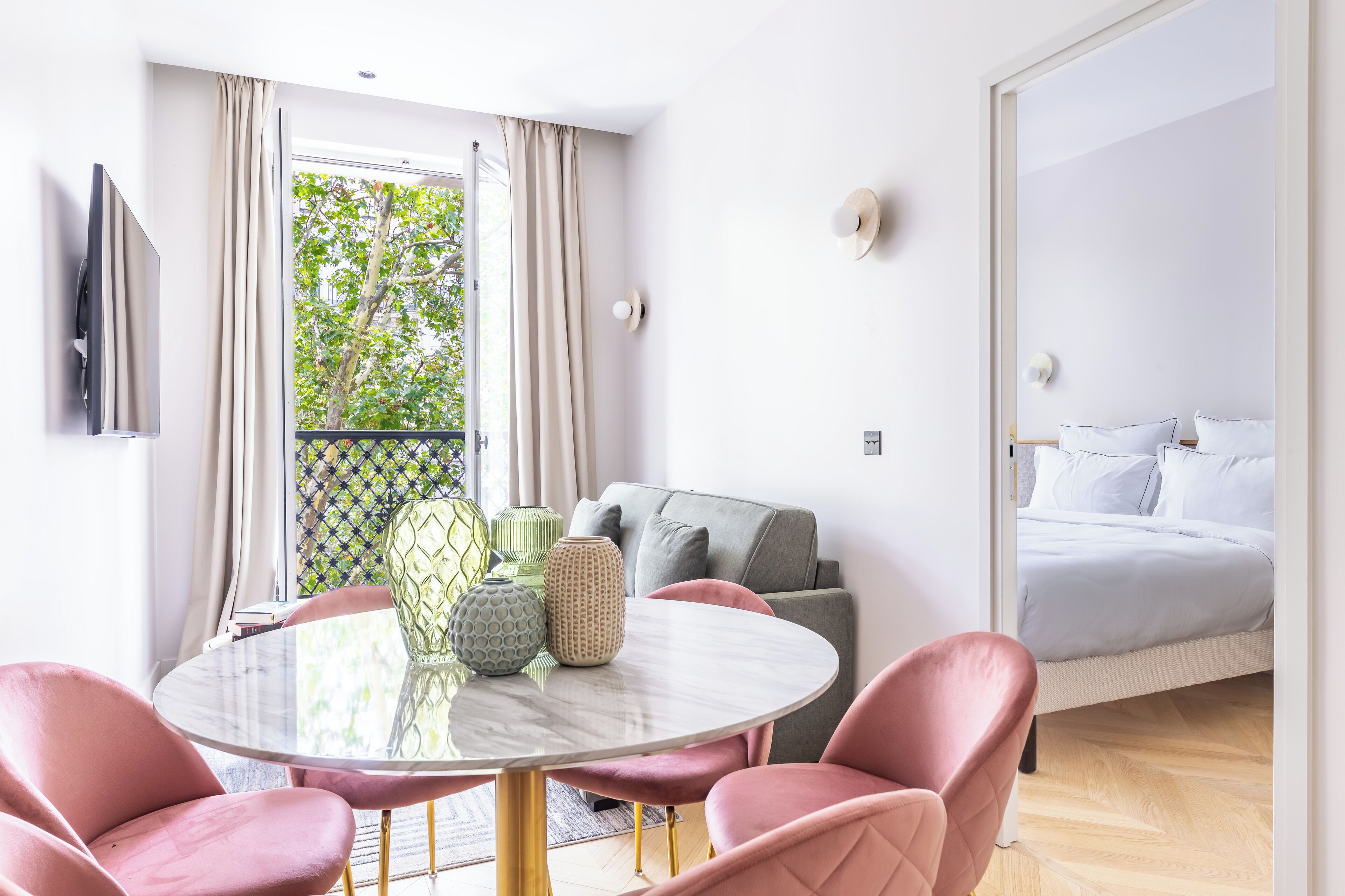 Quarto moderno com mesa redonda, cadeiras cor-de-rosa, sofá cinza e cama com roupa de cama branca.
