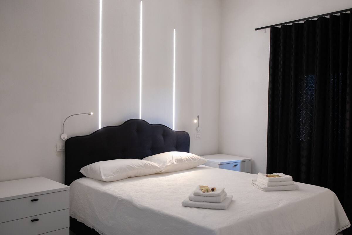 Uma cama bem arrumada com lençóis brancos e duas almofadas, uma mesa de cabeceira com abajur e uma janela com cortinas pretas.