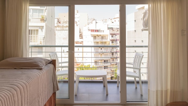 Quarto com cama, varanda com mesa e cadeiras, e vista para a paisagem urbana.