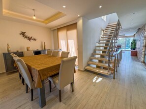 Uma sala de jantar espaçosa com uma mesa comprida e cadeiras, uma escada que leva a um andar superior e vista para o exterior.