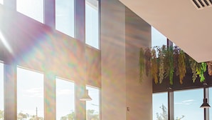 Um espaço interior moderno com grandes janelas, área de refeições com mesas e cadeiras, e plantas penduradas.