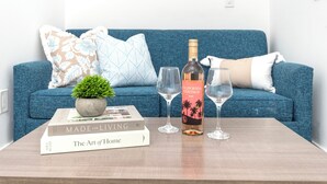 Um sofá azul com almofadas estampadas, uma pequena planta em vaso, duas taças de vinho e uma garrafa de vinho sobre uma mesa de madeira.