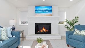 Uma sala de estar moderna com lareira, uma TV de tela plana, dois sofás azuis e um tabuleiro decorativo com velas e uma planta.