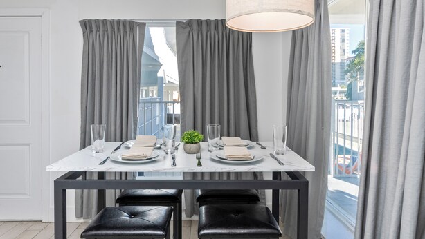 Uma sala de jantar com uma mesa posta para dois, cortinas cinzas, um vaso com planta e vista para edifícios pela janela.